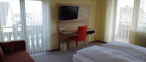 Quarto de hotel com uma cama, uma escrivaninha com cadeira, uma televisão na parede e uma janela com cortinas.