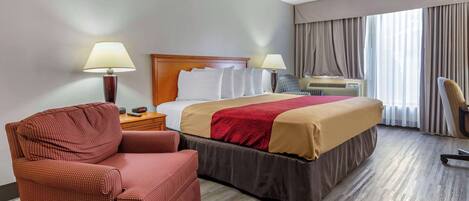 Quarto de hotel com uma cama, uma poltrona vermelha, uma mesa com abajur e uma janela com cortinas.