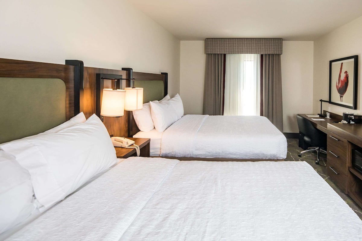 Quarto de hotel com duas camas, uma escrivaninha, uma cadeira, um telefone e uma janela com cortinas.