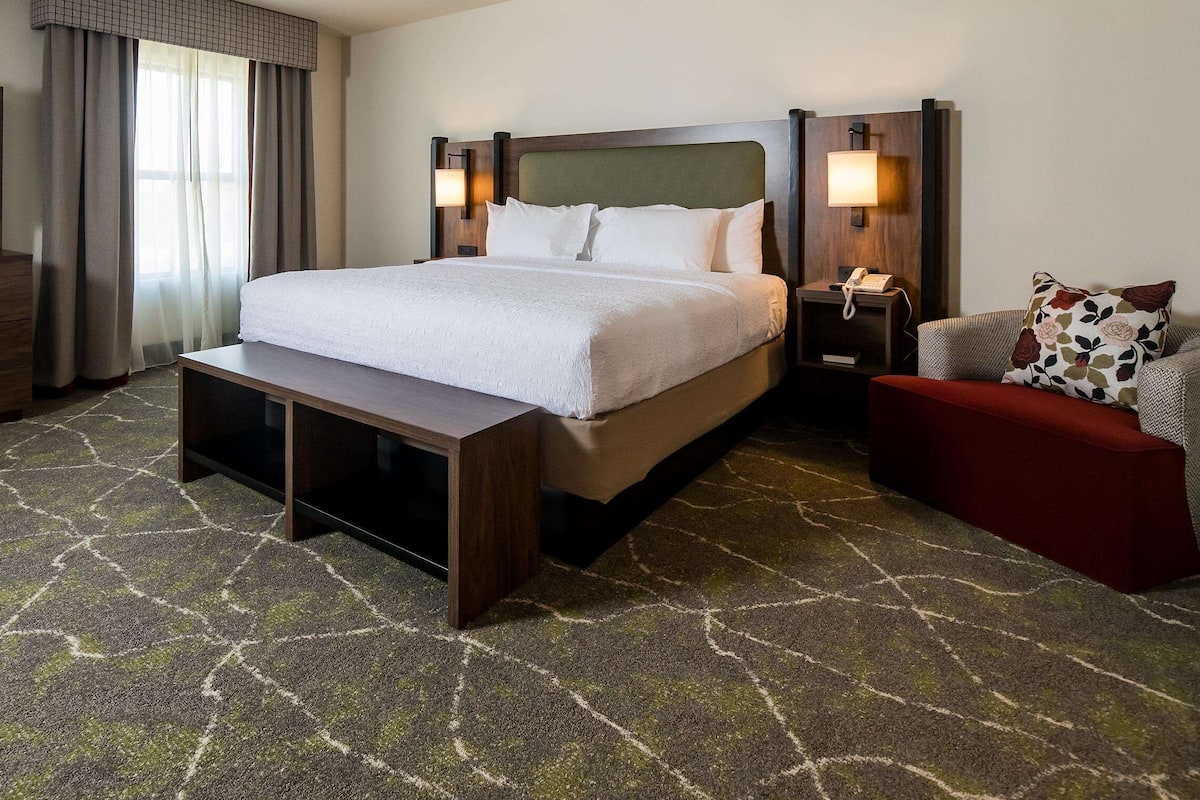 Quarto de hotel com uma cama grande, mesas de cabeceira com luminárias, um pequeno sofá e um banco.