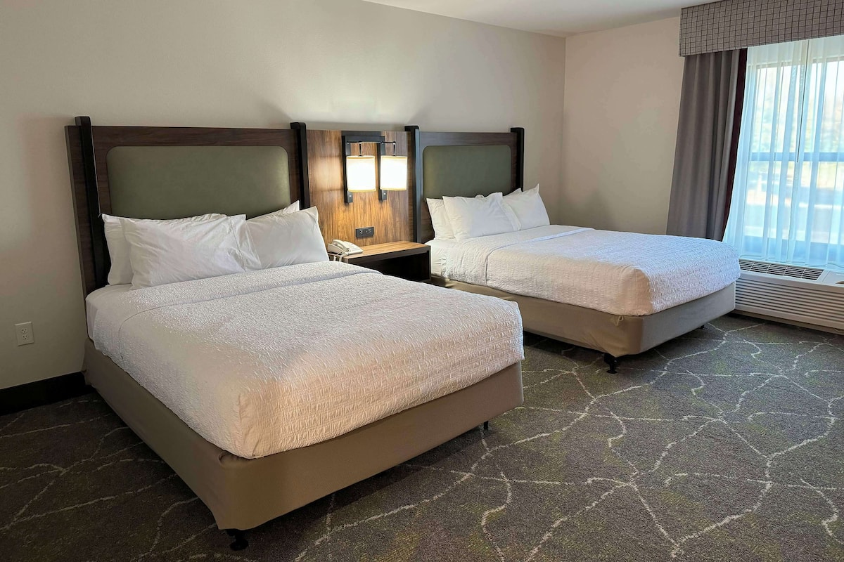 Quarto de hotel com duas camas, mesa de cabeceira, luminária e janela com cortinas.