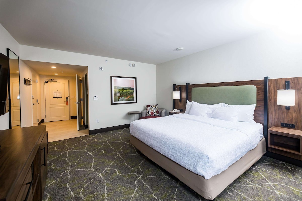 Quarto de hotel com uma cama grande, uma escrivaninha, uma cadeira, uma mesinha de cabeceira e uma luminária fixada na parede.