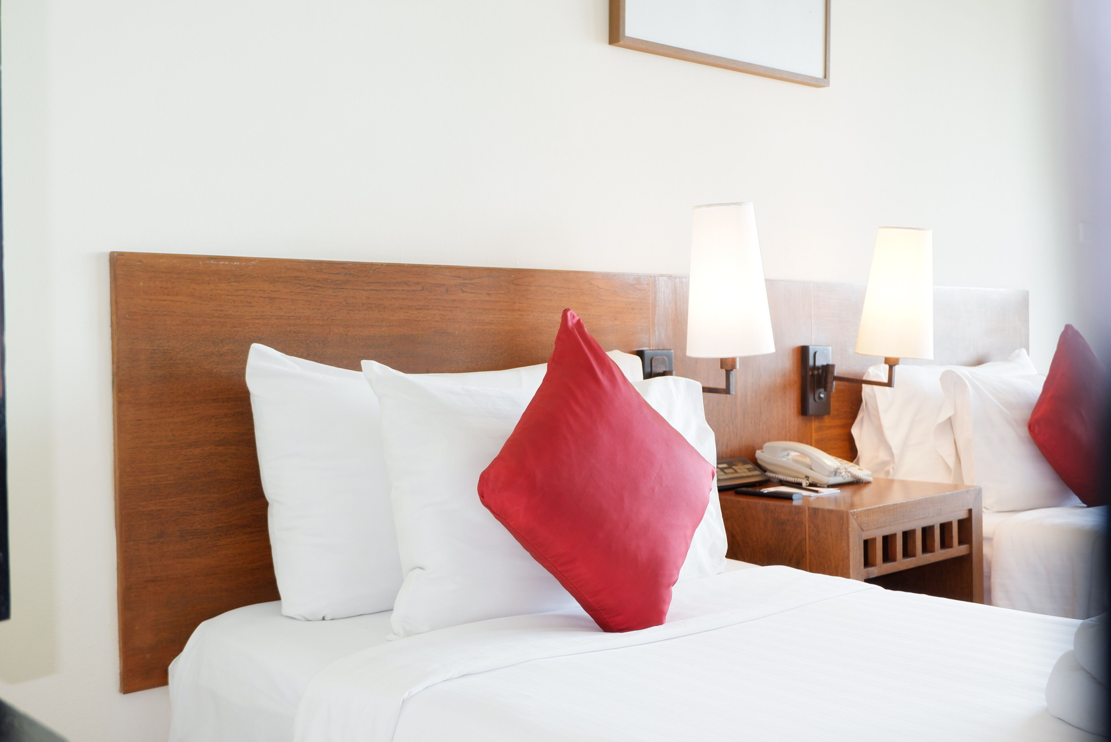 Cama bem arrumada com almofadas brancas e vermelhas, cabeceira de madeira e abajures laterais.