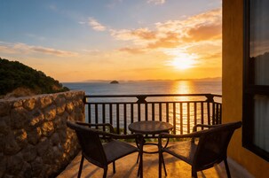 Varanda com vista para o pôr do sol sobre o mar, duas cadeiras de vime e uma parede de pedra.