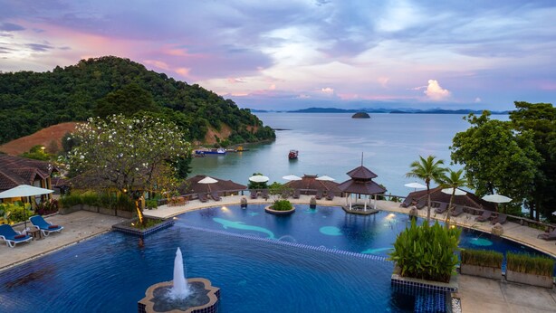 Uma piscina de resort com uma fonte, espreguiçadeiras, guarda-sóis e vista para o mar e colinas.