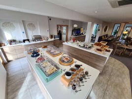 Espaço de café da manhã de hotel com buffet, incluindo diversos alimentos e acompanhamentos.