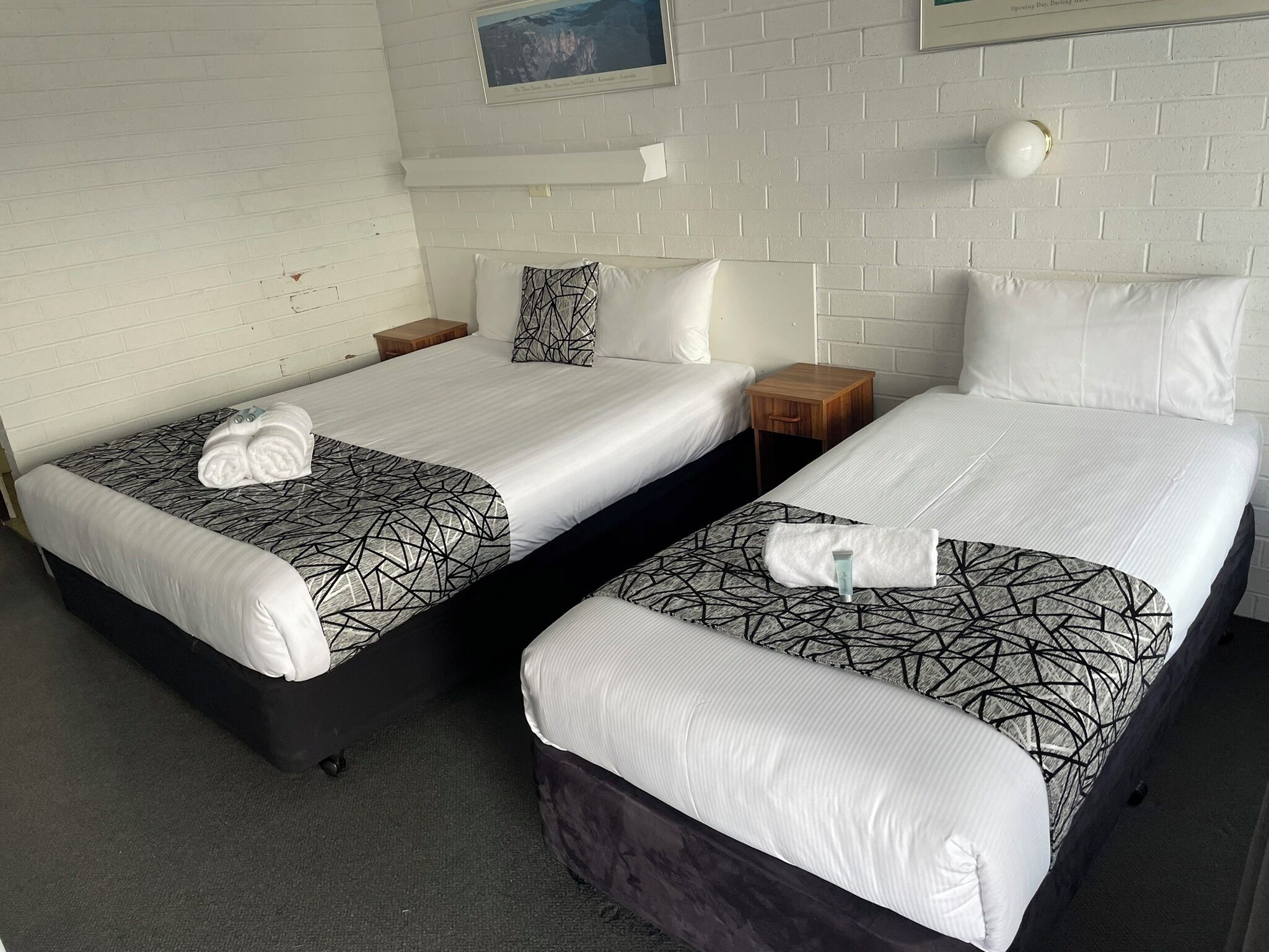 Quarto de hotel com duas camas, cada uma com uma colcha estampada e uma toalha enrolada. Há mesinhas de cabeceira de madeira e uma parede de tijolos brancos com um quadro emoldurado.