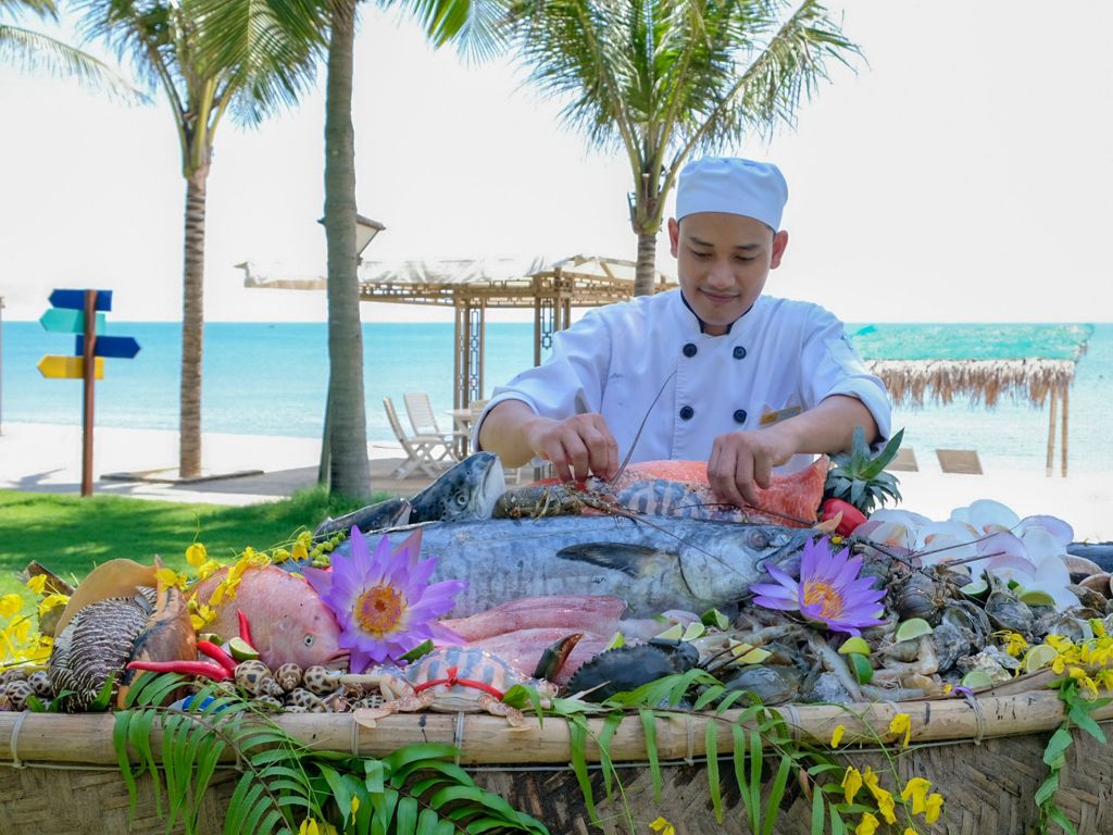 Um chef prepara frutos do mar numa praia com palmeiras e vista clara do oceano.