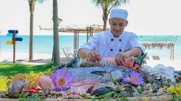 Um chef prepara frutos do mar numa praia com palmeiras e vista clara do oceano.