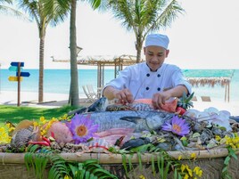Um chef prepara frutos do mar numa praia com palmeiras e vista clara do oceano.