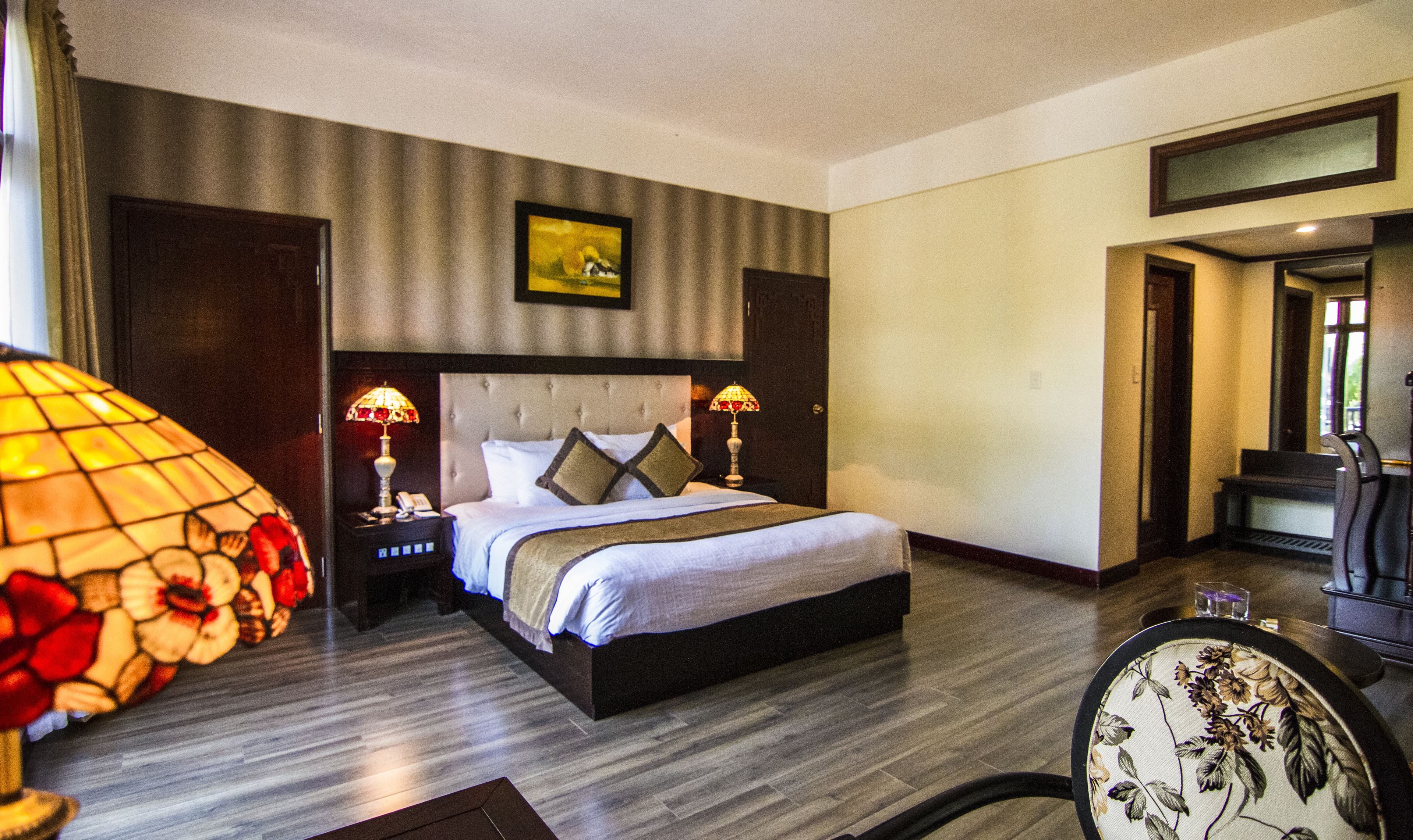 Quarto de hotel com uma cama grande, duas luminárias de cabeceira, uma mesinha de cabeceira, um espelho e um quadro na parede.
