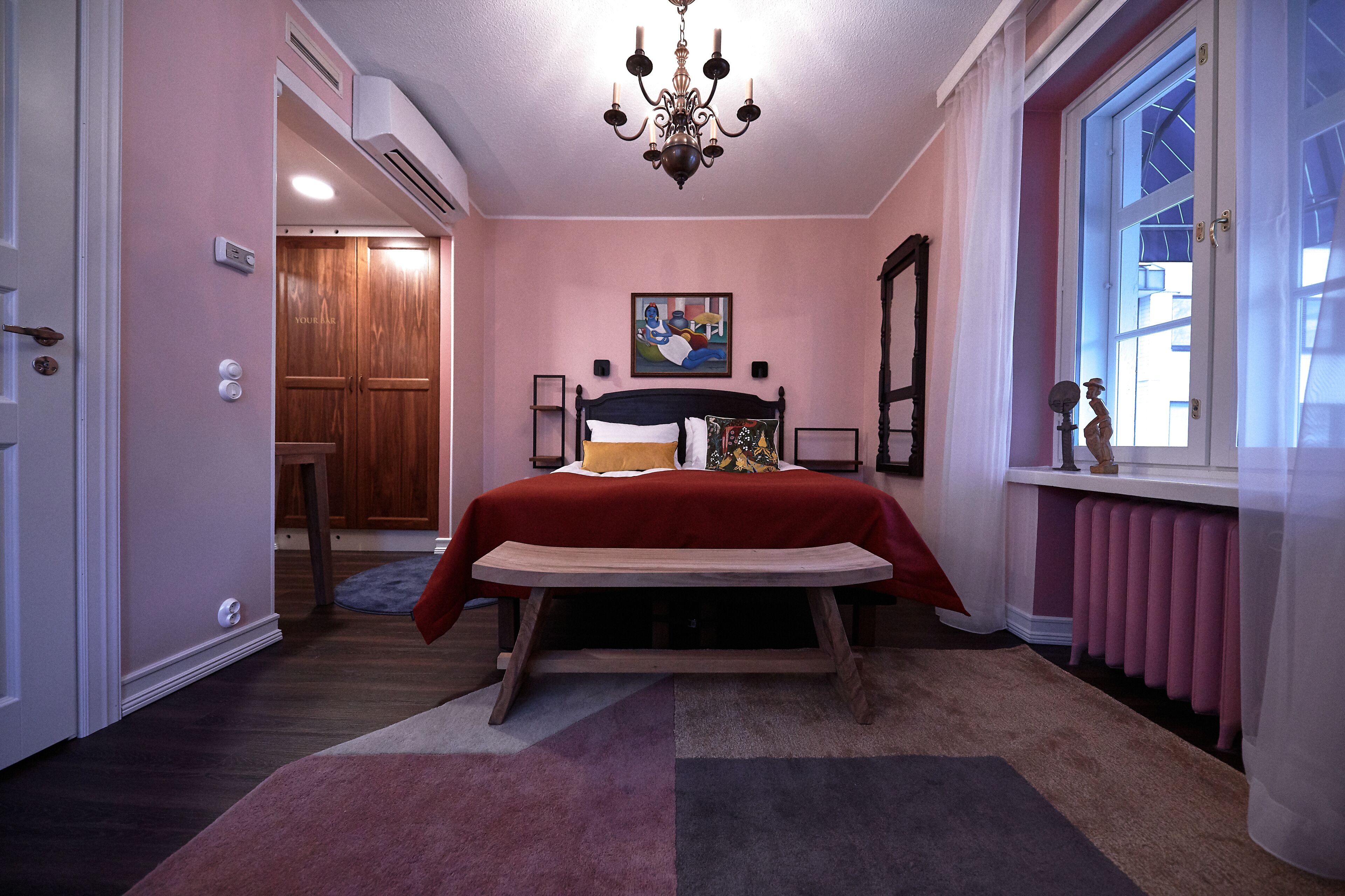 Quarto com cama, um lustre, janela com cortinas, radiador e porta.