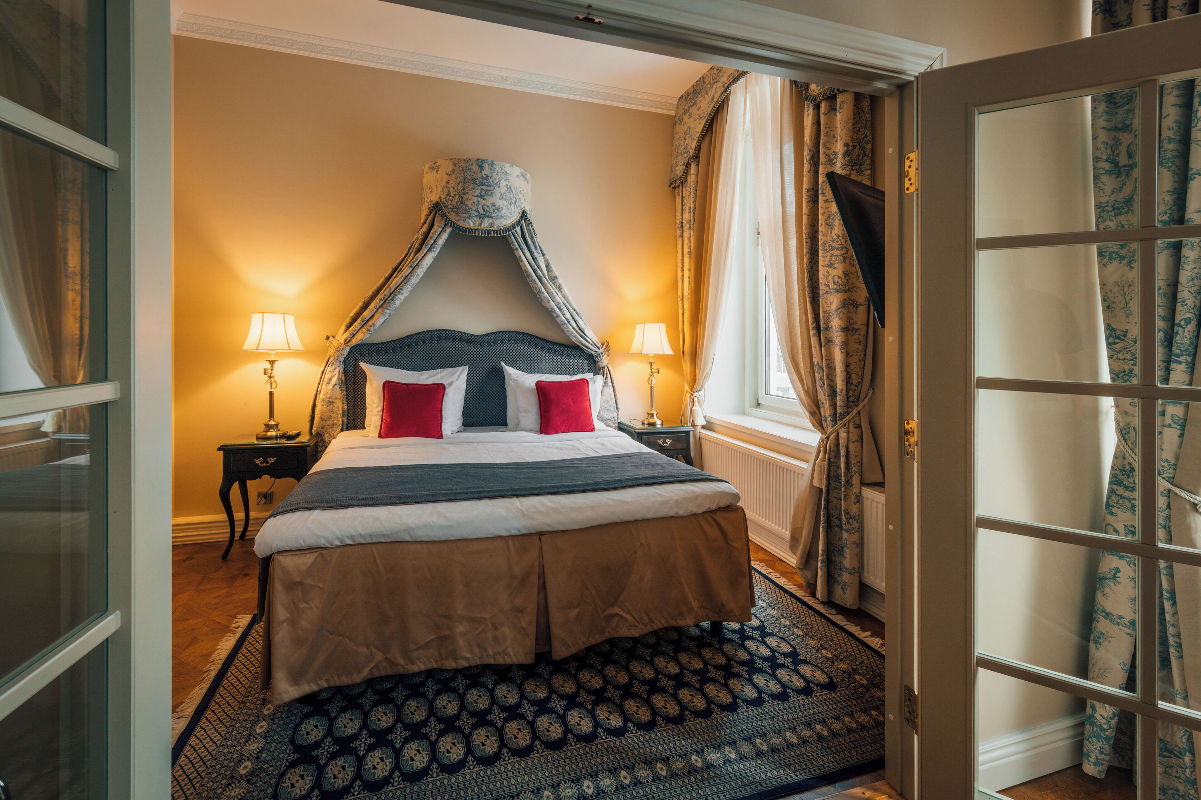 Quarto com cama de dossel, duas luminárias de cabeceira, tapete estampado e vista através de uma porta aberta.