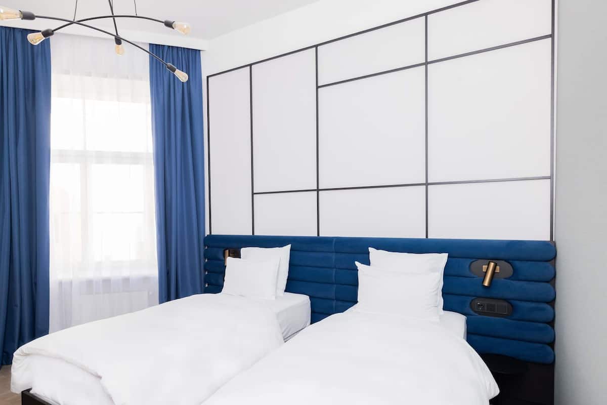 Quarto de hotel com duas camas, uma janela grande com cortinas azuis e um design moderno na parede.