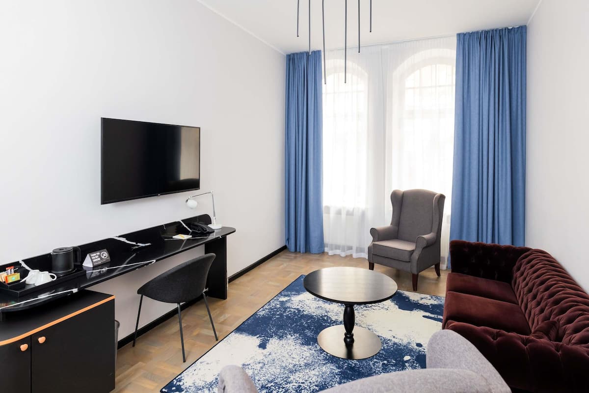 Quarto de hotel moderno com televisão de tela plana, escrivaninha com cadeira, mesa redonda, sofá e uma grande janela com cortinas.