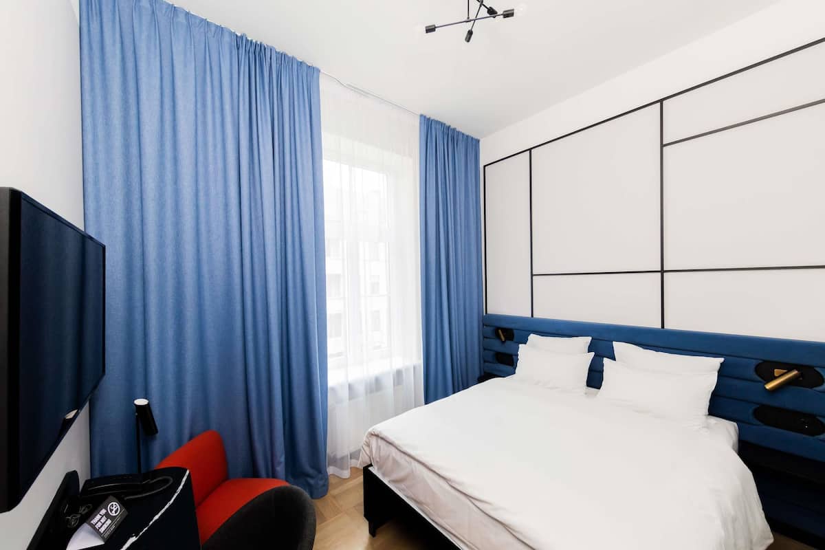 Quarto de hotel com cama, televisão, cadeira e janela com cortinas.