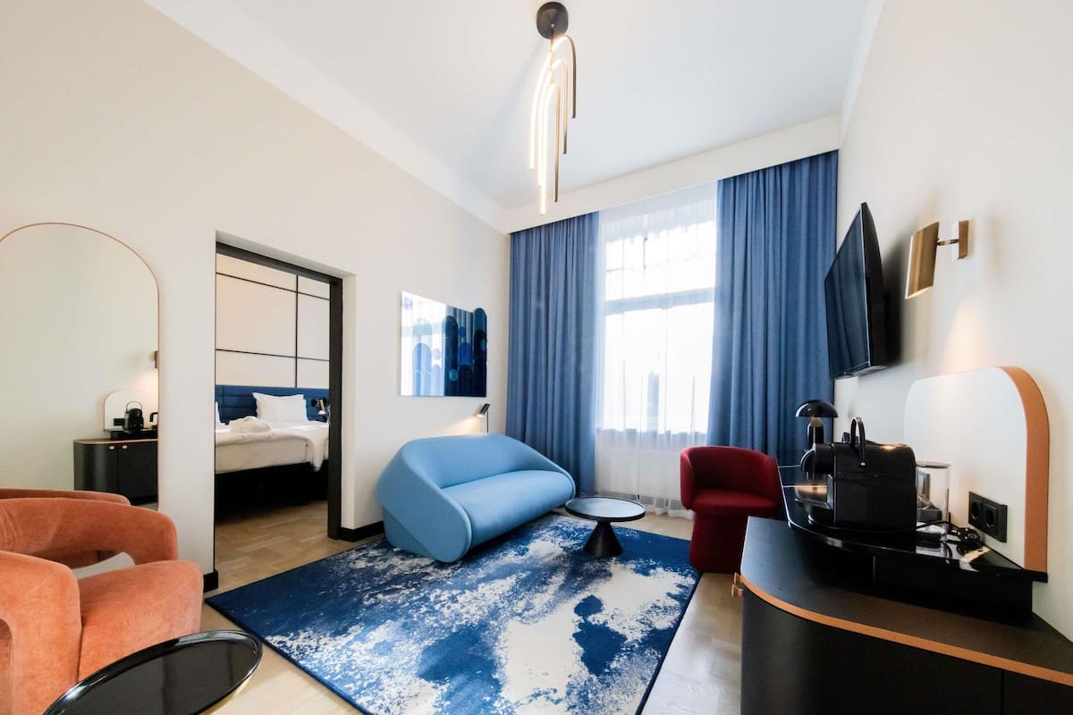 Quarto de hotel moderno com uma cama, um sofá azul, uma poltrona vermelha, uma mesa redonda pequena e uma área de cozinha compacta.