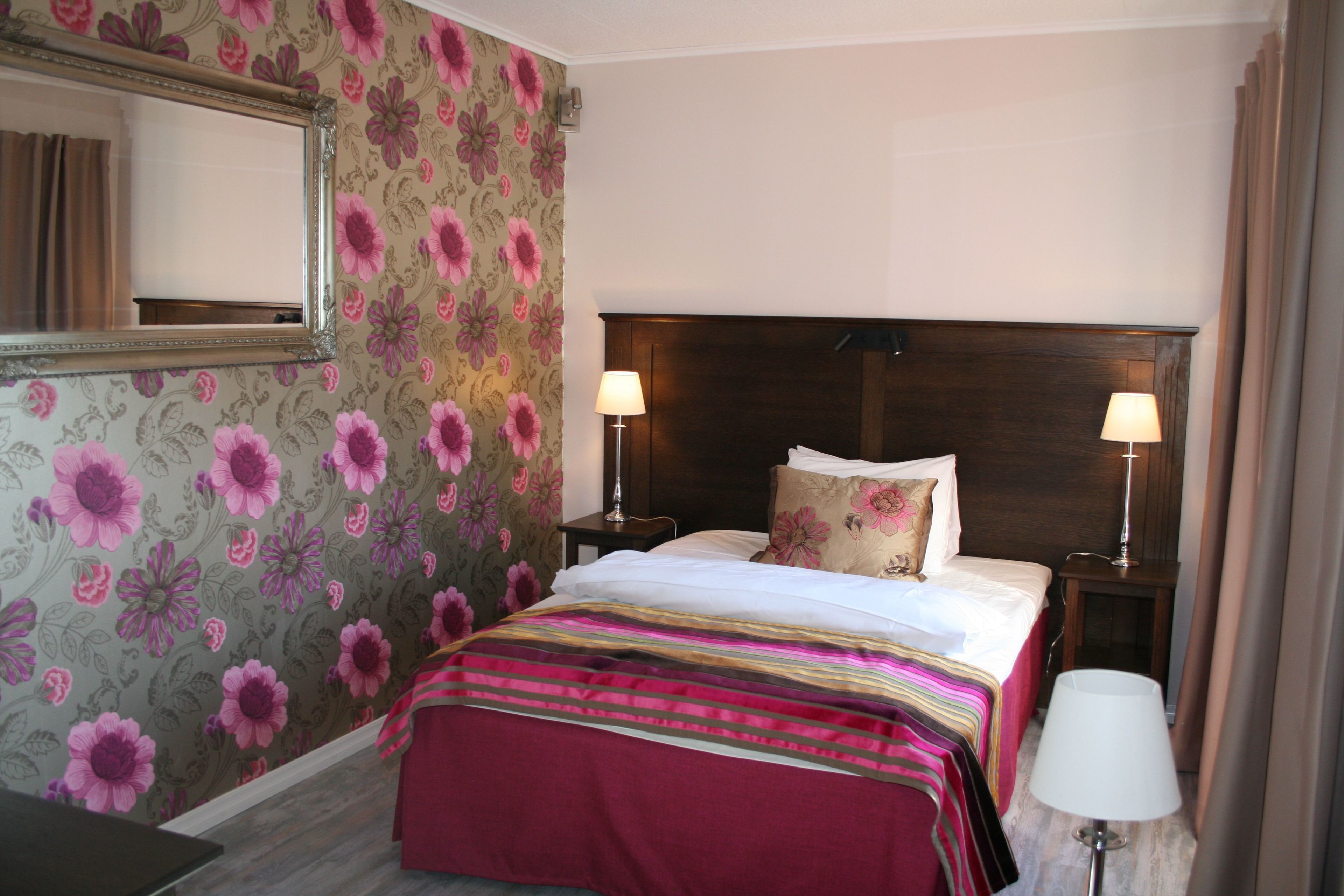 Quarto de hotel com uma cama grande, papel de parede floral, duas mesas de cabeceira com luminárias e um espelho na parede.