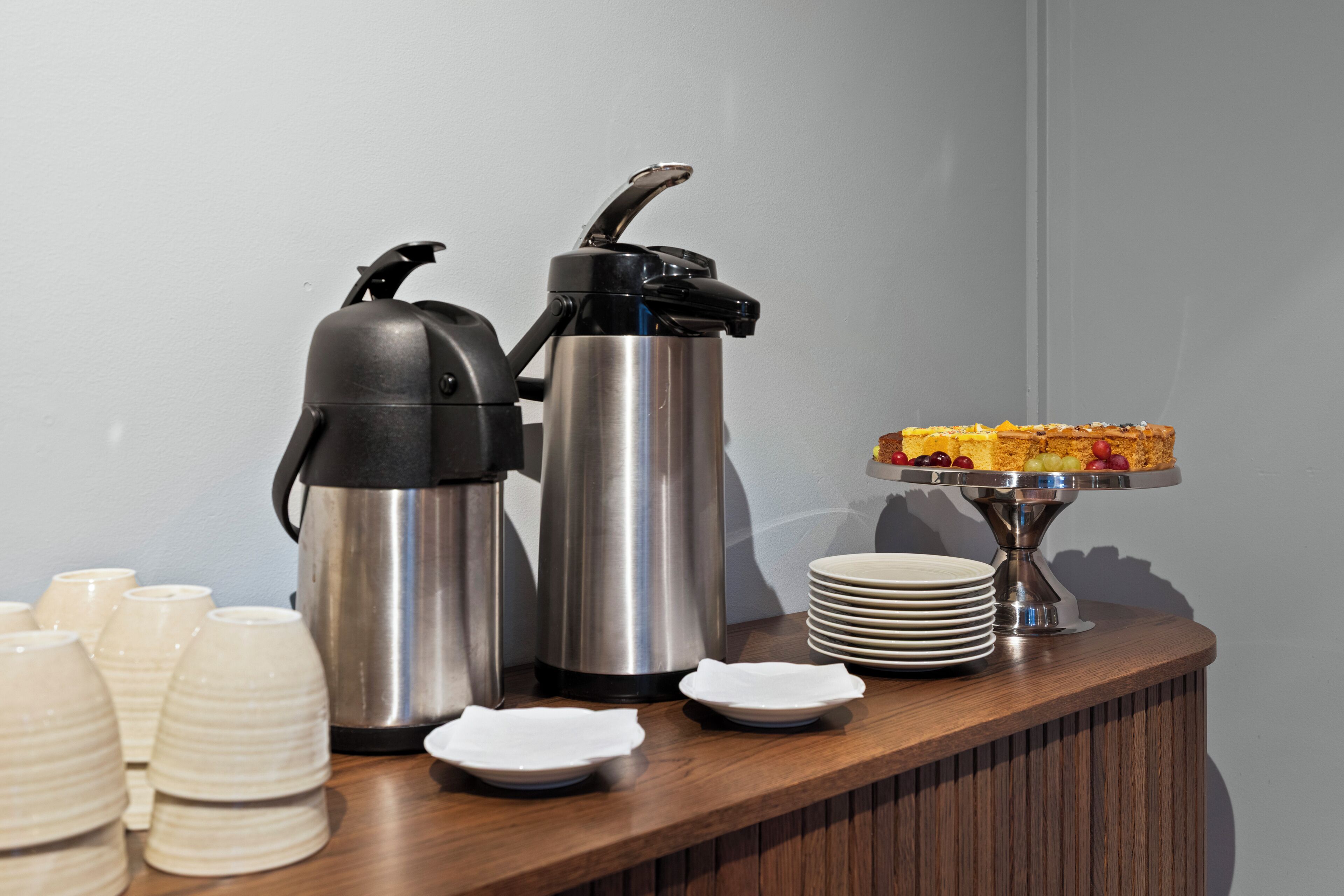 Mesa de madeira com buffet, incluindo máquina de café, pratos e um bolo.