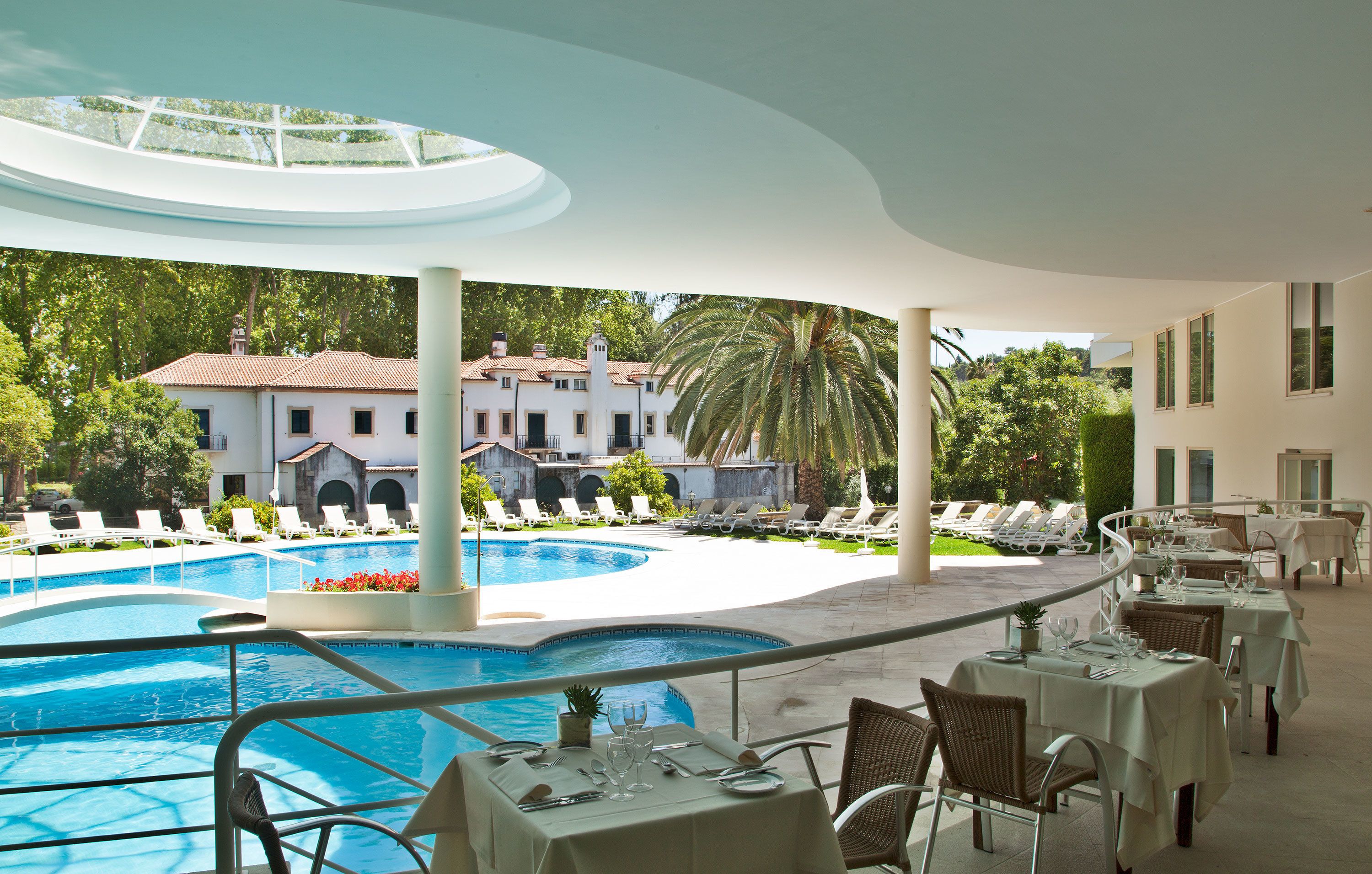 Um restaurante à beira da piscina com mesas e cadeiras, uma piscina e vista para um edifício com telhado em azulejos.