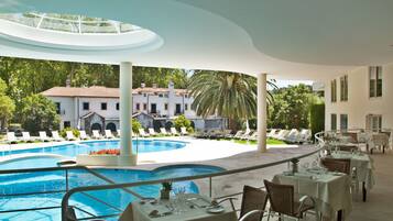 Um restaurante à beira da piscina com mesas e cadeiras, uma piscina e vista para um edifício com telhado em azulejos.