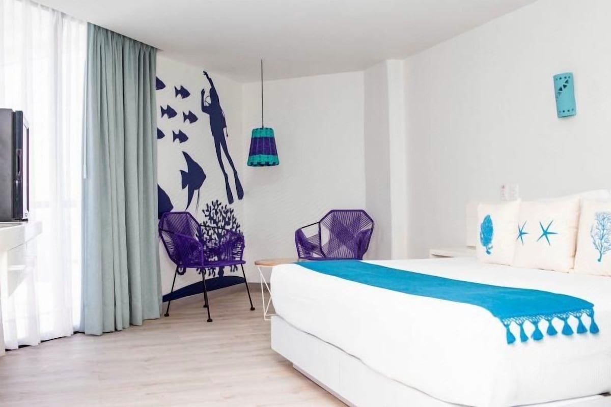 Quarto de hotel com cama, televisão, painel decorativo na parede e duas cadeiras roxas.