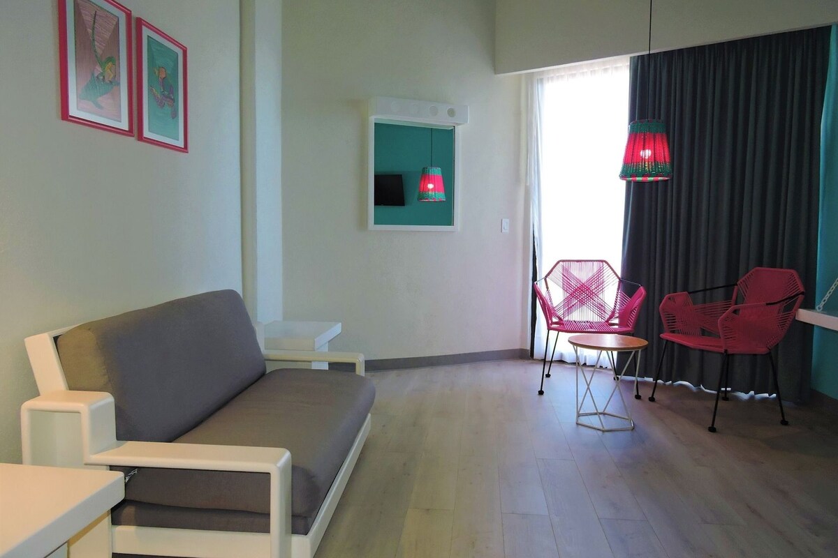 Uma sala de estar moderna com um sofá, uma mesinha, duas cadeiras vermelhas, um espelho com moldura verde-água e uma luminária pendente vermelha.