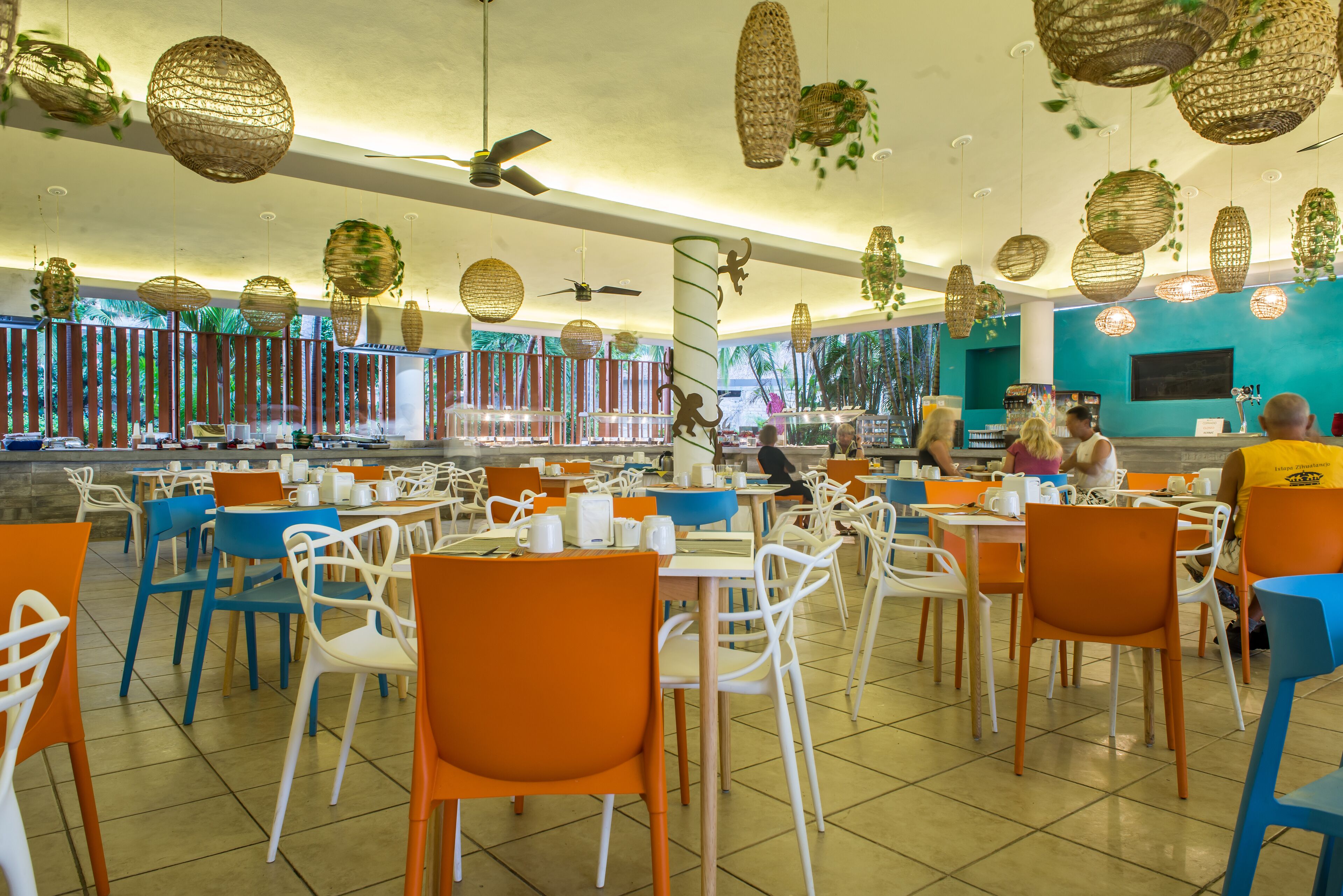 Uma sala de jantar espaçosa com pé-direito alto, luminárias de vime suspensas e diversas opções de assento, incluindo cadeiras laranja e brancas.