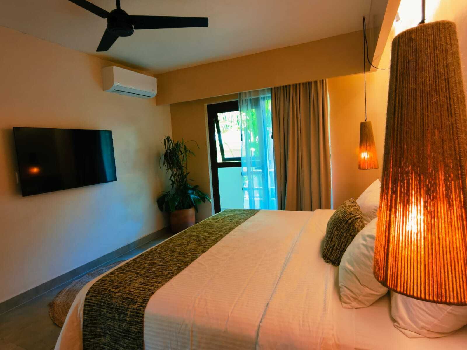 Quarto de hotel com cama, ventoinha no teto, aparelho de ar condicionado, televisão, planta em vaso, janela com cortinas e luminária pendente.
