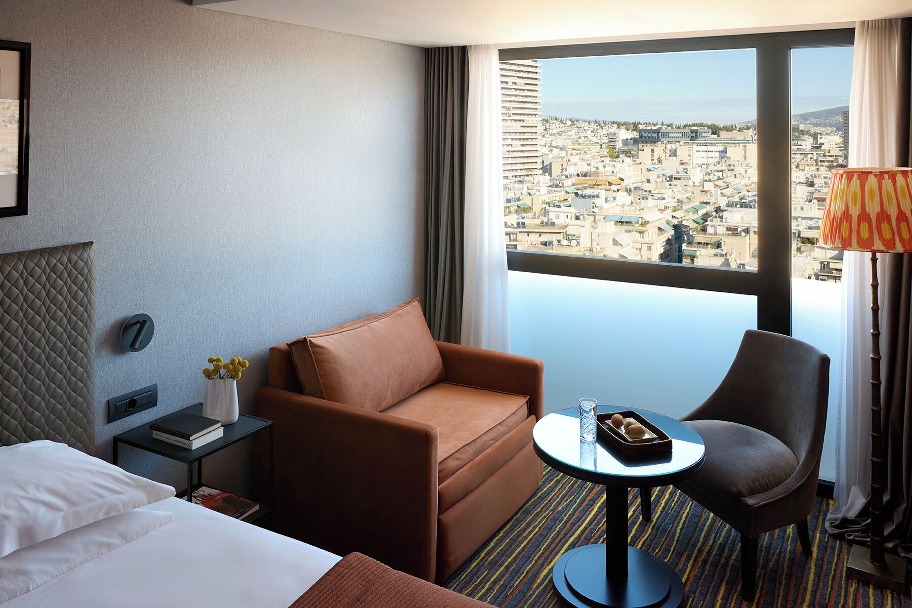 Quarto de hotel com cama, poltrona marrom, mesa redonda pequena e vista para a cidade através de uma grande janela.