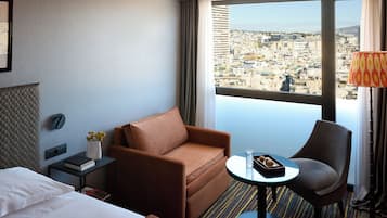 Quarto de hotel com cama, poltrona marrom, mesa redonda pequena e vista para a cidade através de uma grande janela.