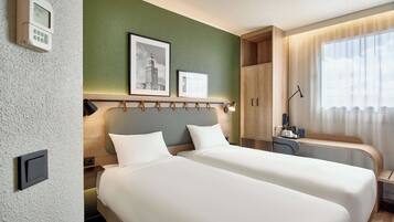 Quarto de hotel com duas camas, uma mesinha de cabeceira, abajur, quadro emoldurado e janela com cortinas.