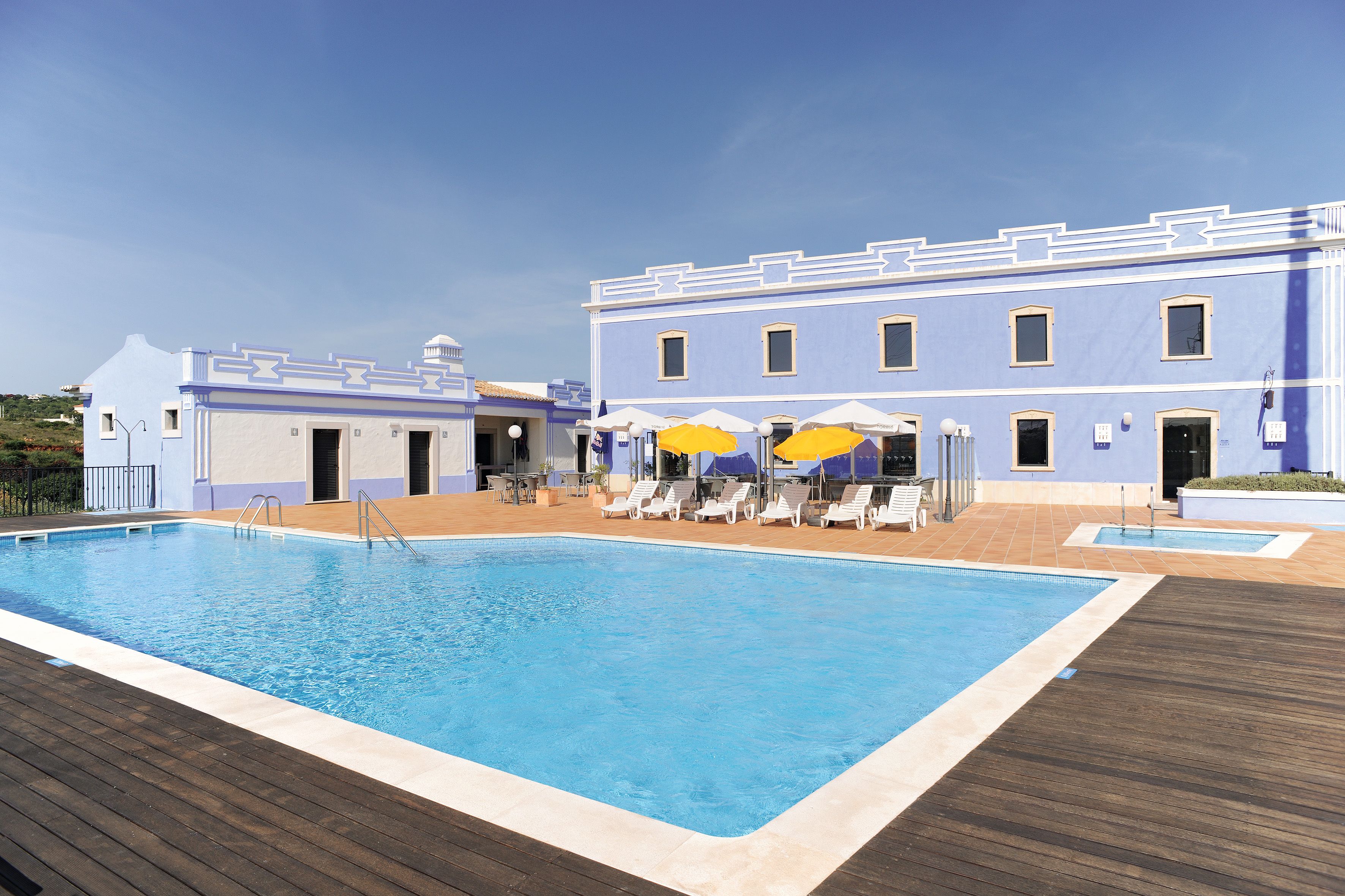 Piscina com espreguiçadeiras e guarda-sóis, um deck de madeira e um edifício com fachada azul.