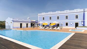 Piscina com espreguiçadeiras e guarda-sóis, um deck de madeira e um edifício com fachada azul.