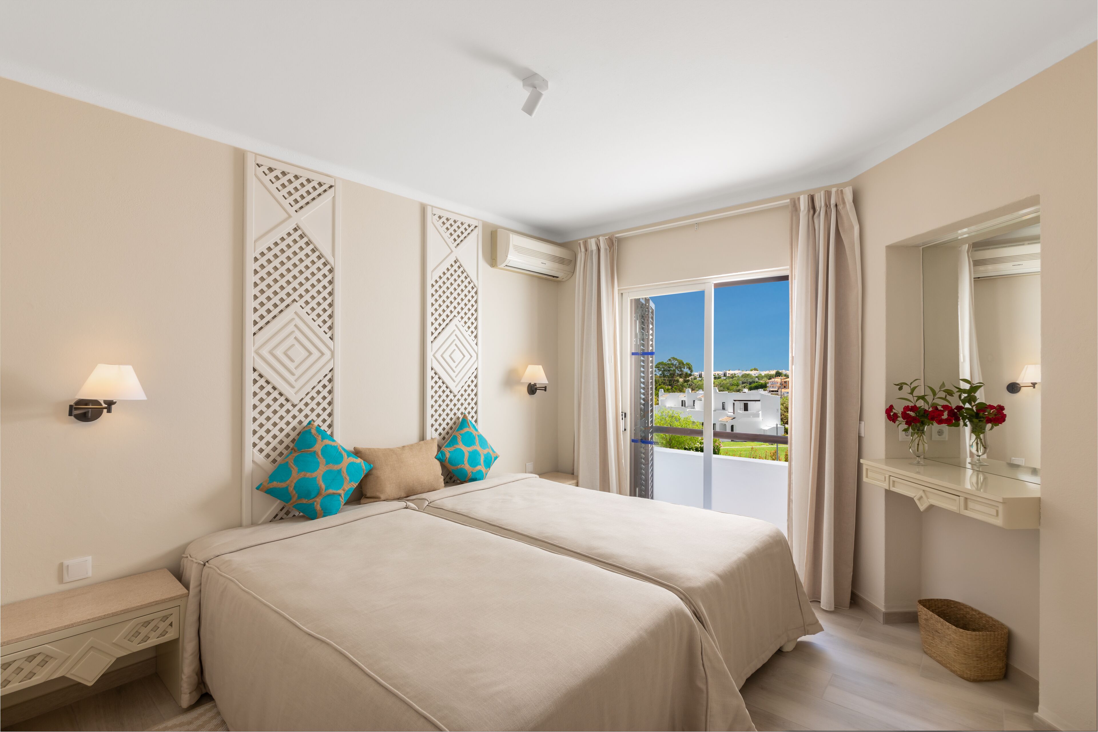 Quarto de hotel com cama, janela com cortinas, ar condicionado fixado na parede, mesa de cabeceira com abajur e uma pequena cesta.