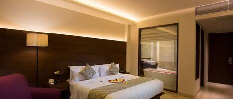 Quarto de hotel com uma cama grande, uma mesa de cabeceira, uma poltrona vermelha, um banheiro com banheira e um espelho.