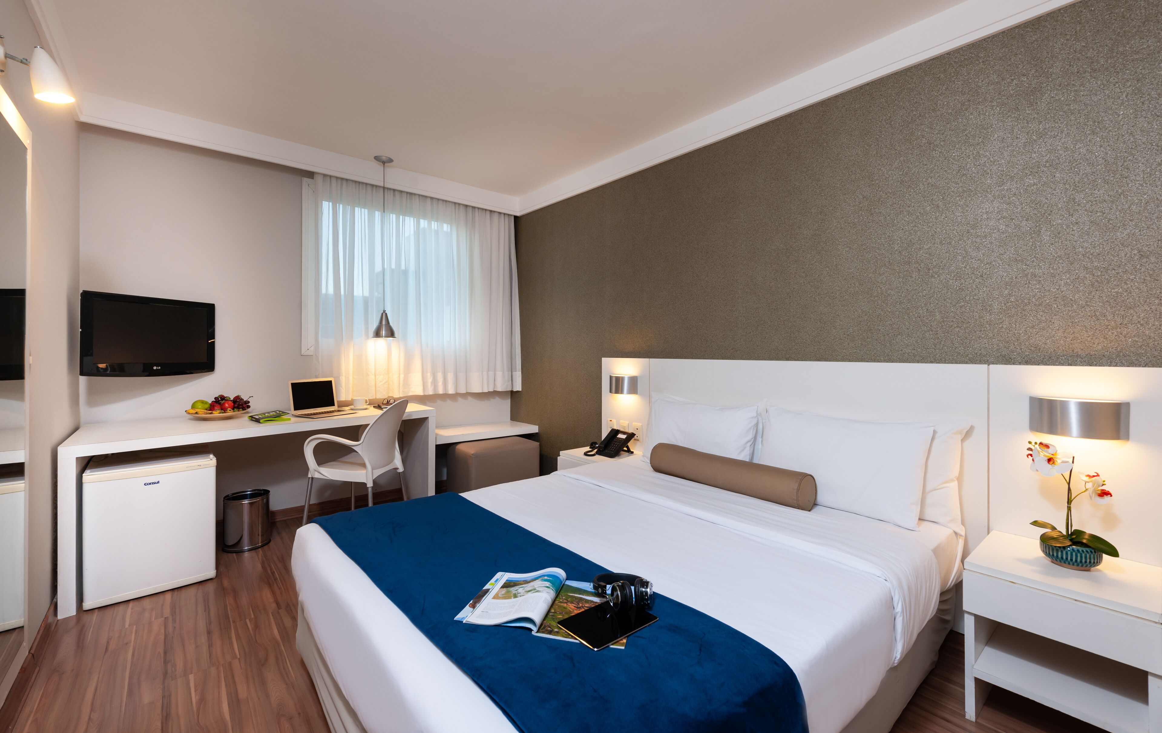 Quarto de hotel com cama, escrivaninha, cadeira e um pequeno frigorífico.