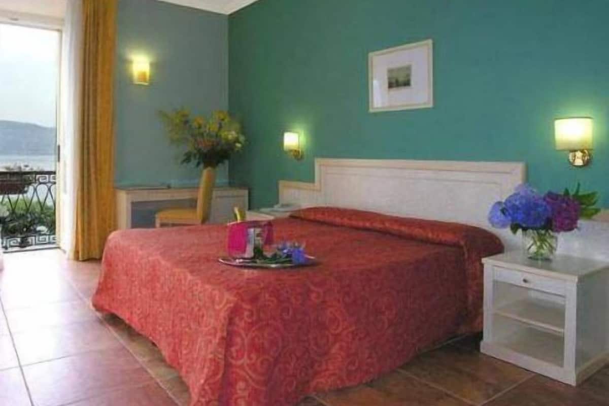 Quarto de hotel com uma cama, uma mesa de cabeceira, um vaso com flores e vista para o exterior.