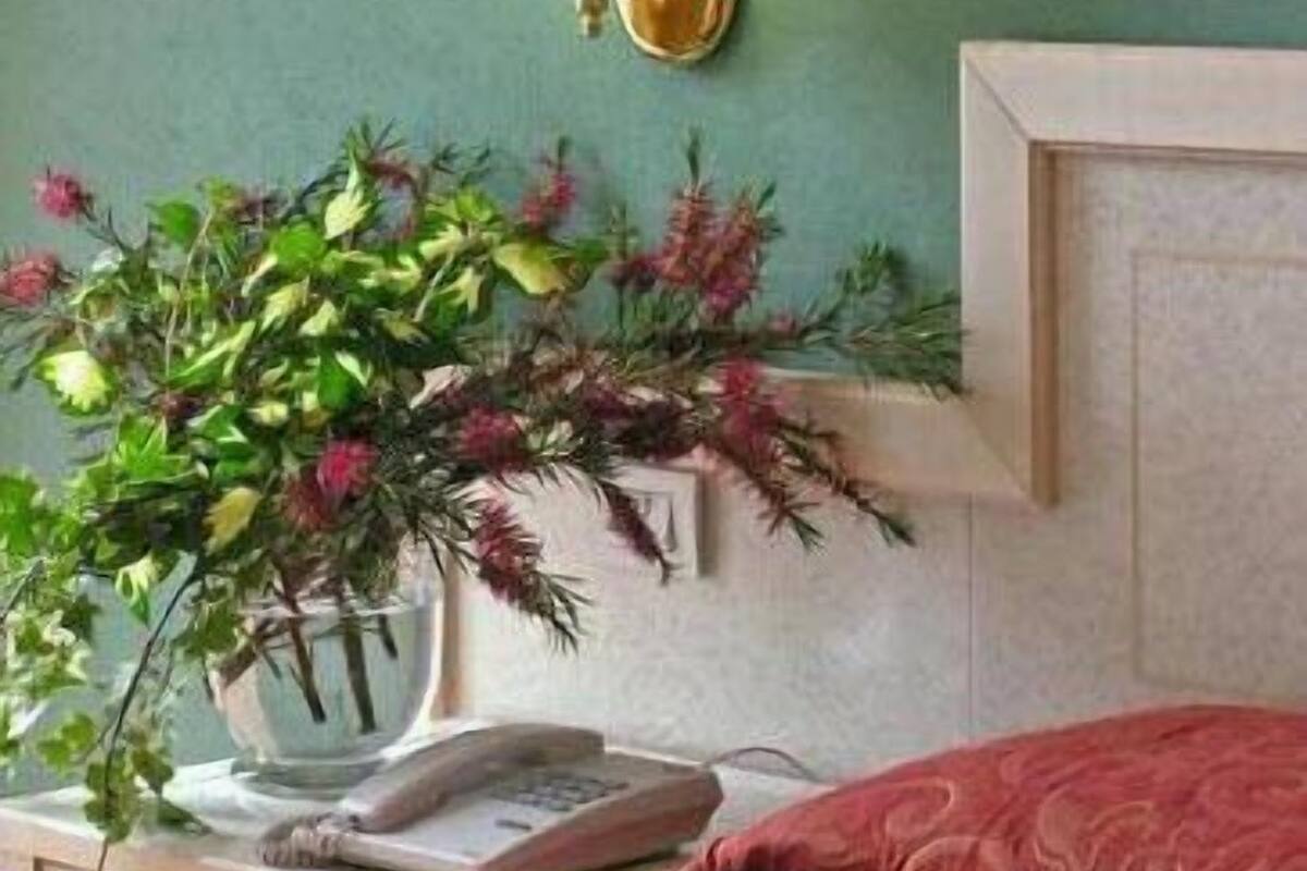 Quarto com cama, mesa de cabeceira com abajur, vaso com flores e um livro.