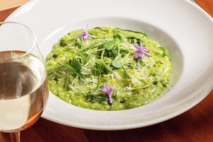 Prato branco com um recipiente verde adornado com flores roxas e ervas, acompanhado de um copo de vinho branco sobre uma mesa de madeira.