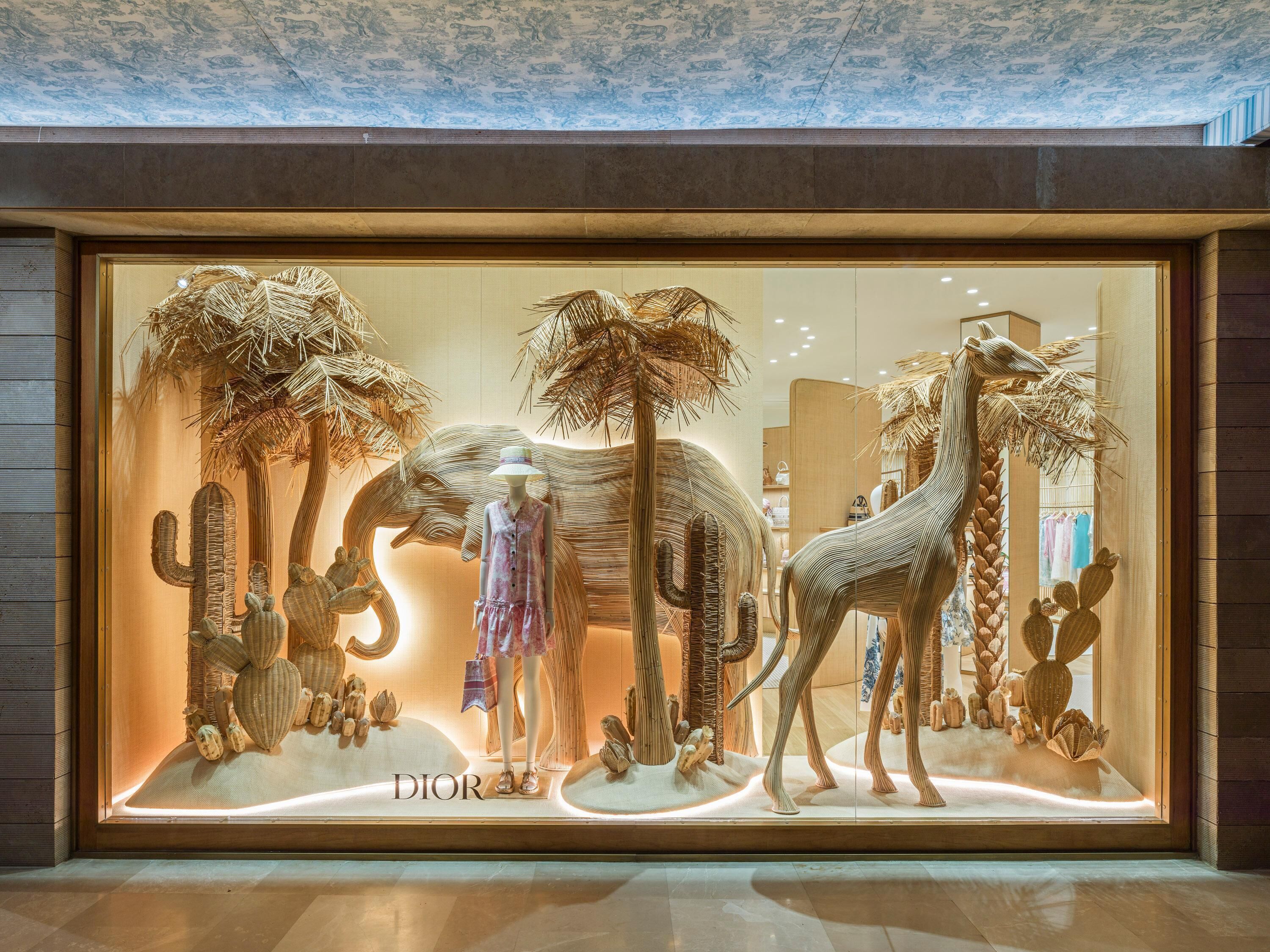 Vitrine da loja Dior com um manequim usando um vestido rosa e um cenário temático de deserto com cactos e palmeiras.