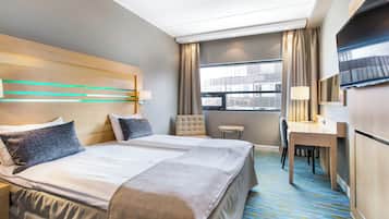 Quarto de hotel com uma cama grande, escrivaninha, cadeira, televisão e janela com cortinas.
