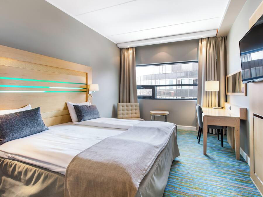 Quarto de hotel com uma cama grande, escrivaninha, cadeira, televisão e janela com cortinas.