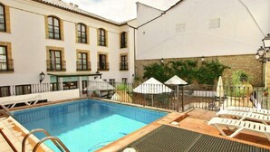 Piscina com deck ladrilhado, rodeada por um edifício branco com várias janelas e varandas.