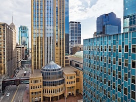 Uma área urbana moderna com edifícios altos, uma estrutura cilíndrica imponente e um edifício azul com padrão de grade.