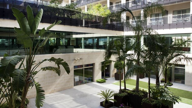 Um edifício com vários andares, varandas, um pátio com plantas em vasos e fachada de vidro.