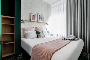 Cama bem arrumada com um cobertor rosa, uma estante de madeira e uma parede com duas fotos emolduradas.