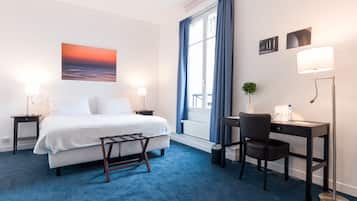 Quarto de hotel com cama, escrivaninha, cadeira, luminária, planta e janela com cortinas.