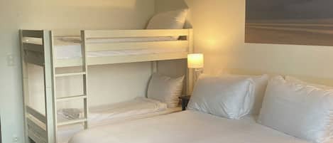 Quarto de hotel com uma cama beliche, uma cama de solteiro, uma escrivaninha e uma mesinha de cabeceira.