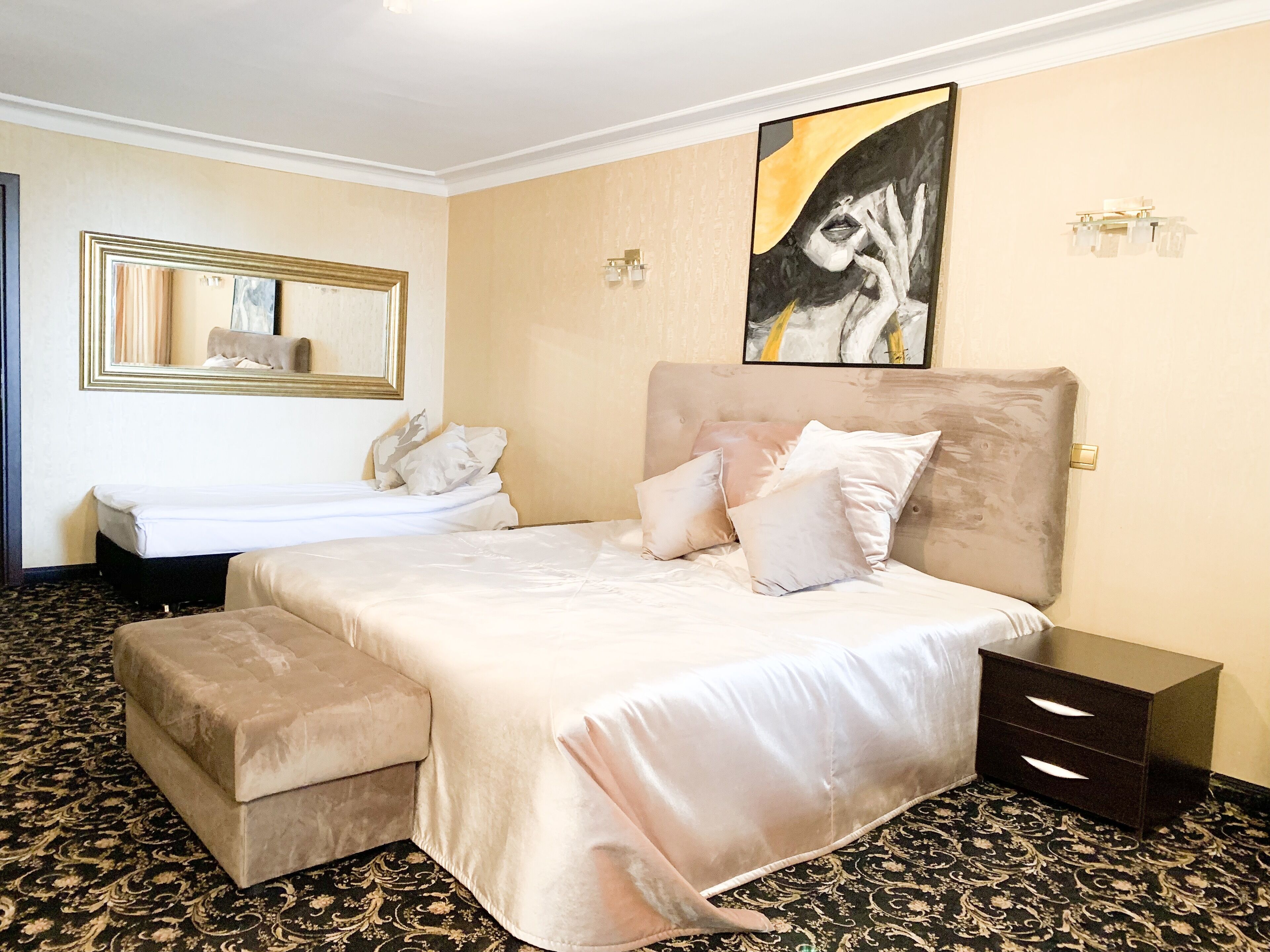 Quarto de hotel com uma cama grande, uma mesa de cabeceira, um banco, um espelho e um quadro na parede.